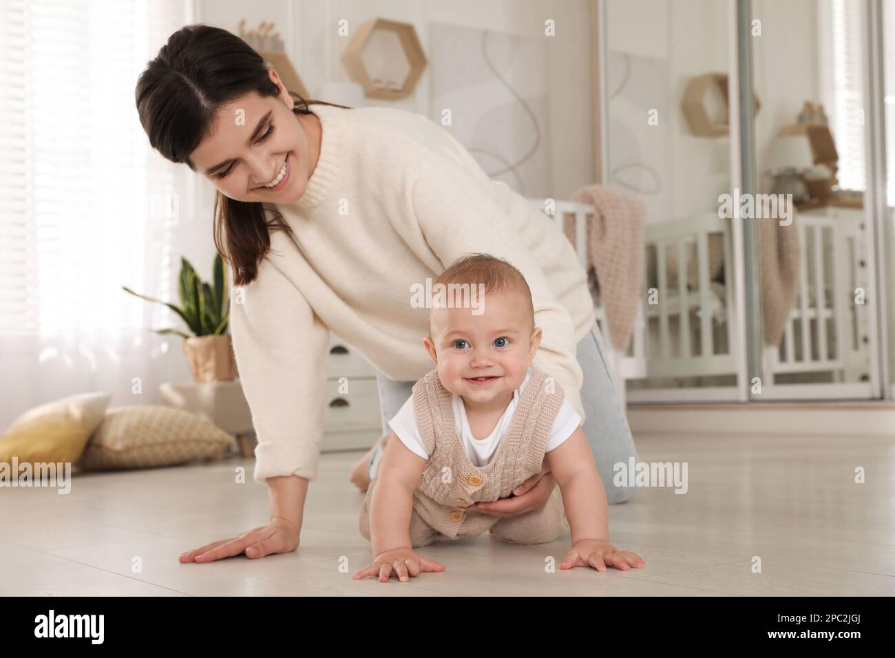Happy young mother helping her cute baby to crawl on floor at home ...