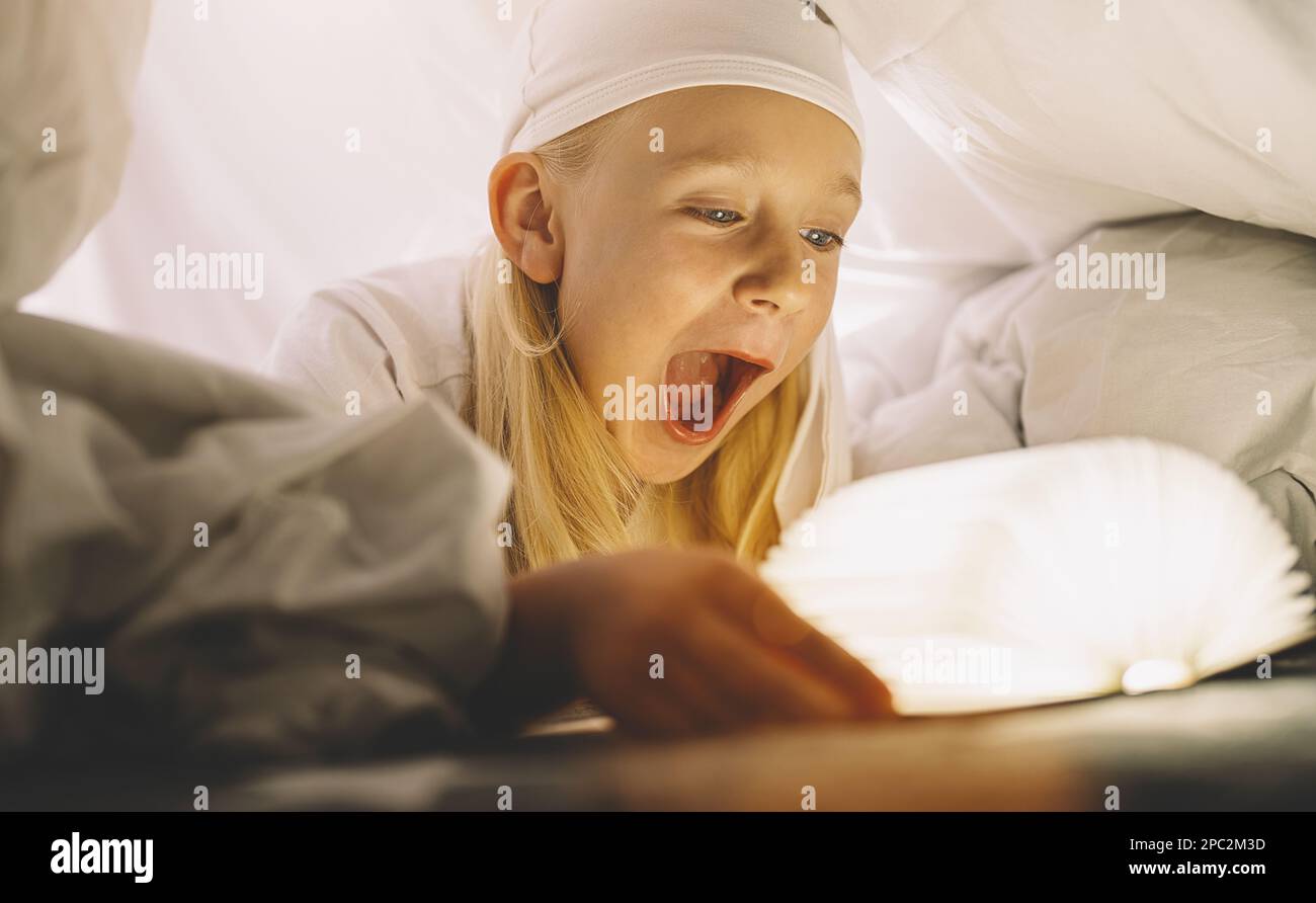 astonished young child reading book under blanket at night. Magic light ...