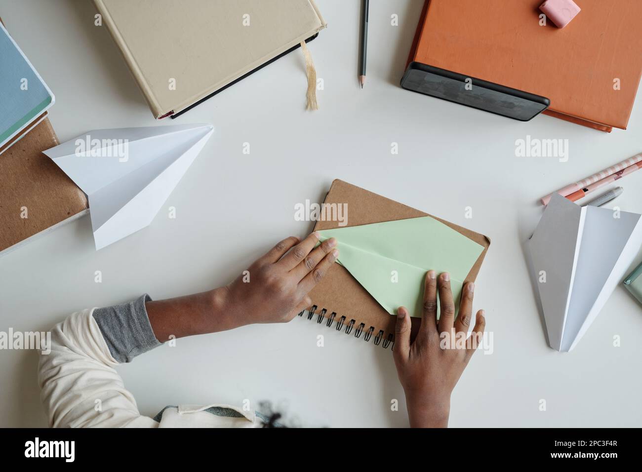 Top view of hands of youthful schoolboy making paper plane by desk with ...