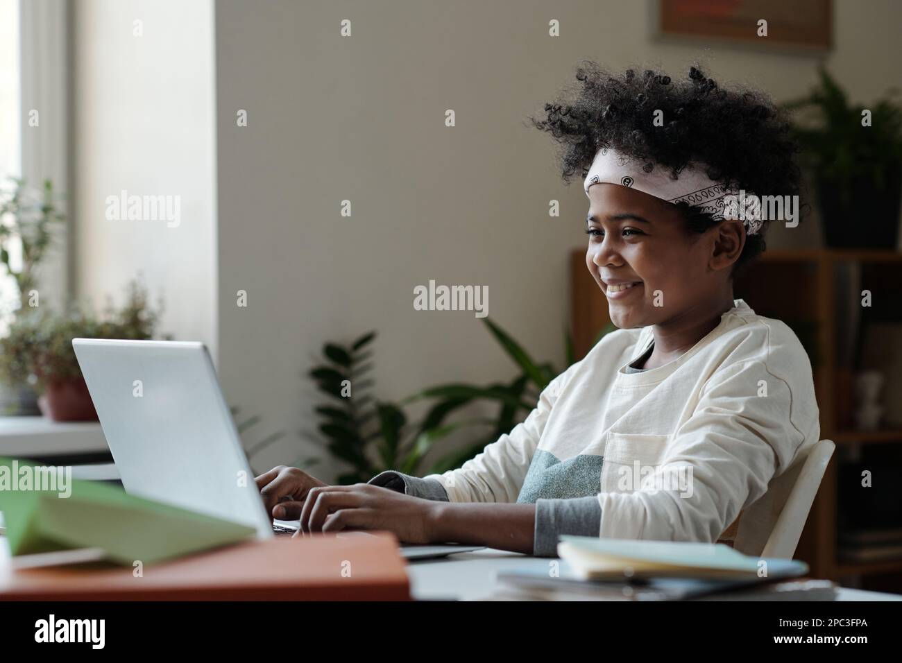 Cute smiling schoolchild sitting by desk in front of laptop and typing ...