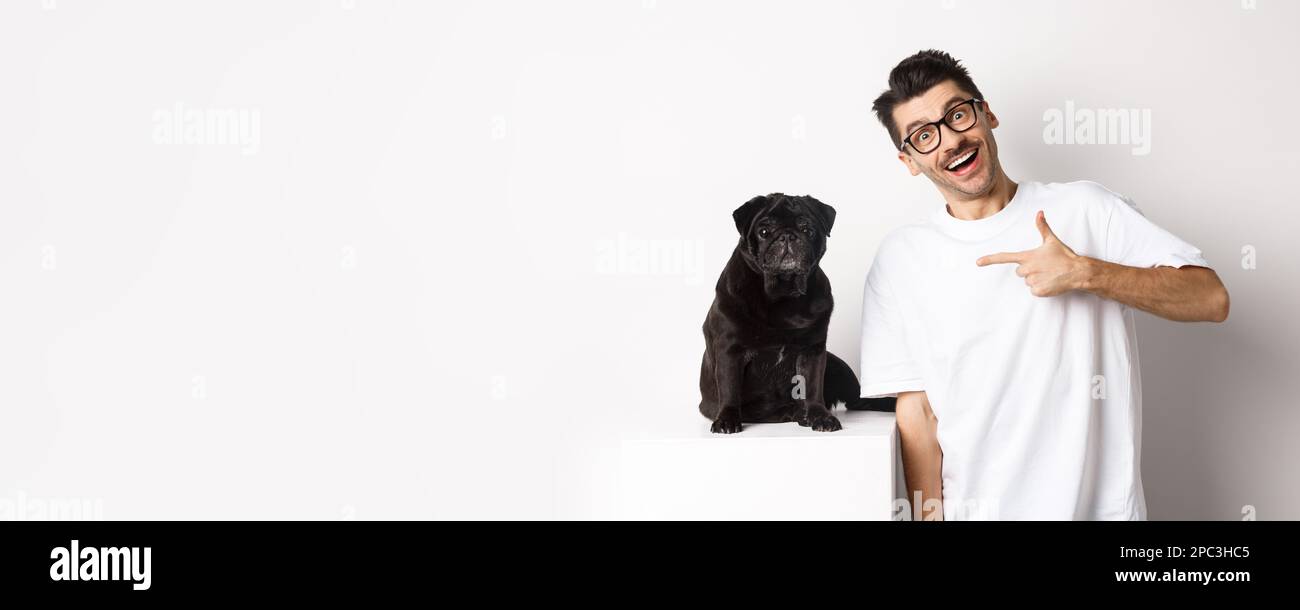 Happy young man showing his cute dog, pointing finger at black pug and ...