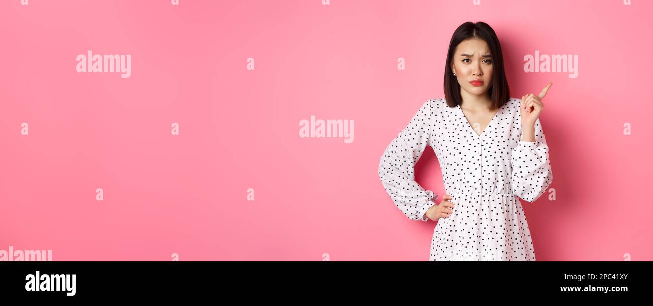 Sad and gloomy asian girl frowning, pointing finger at upper right ...