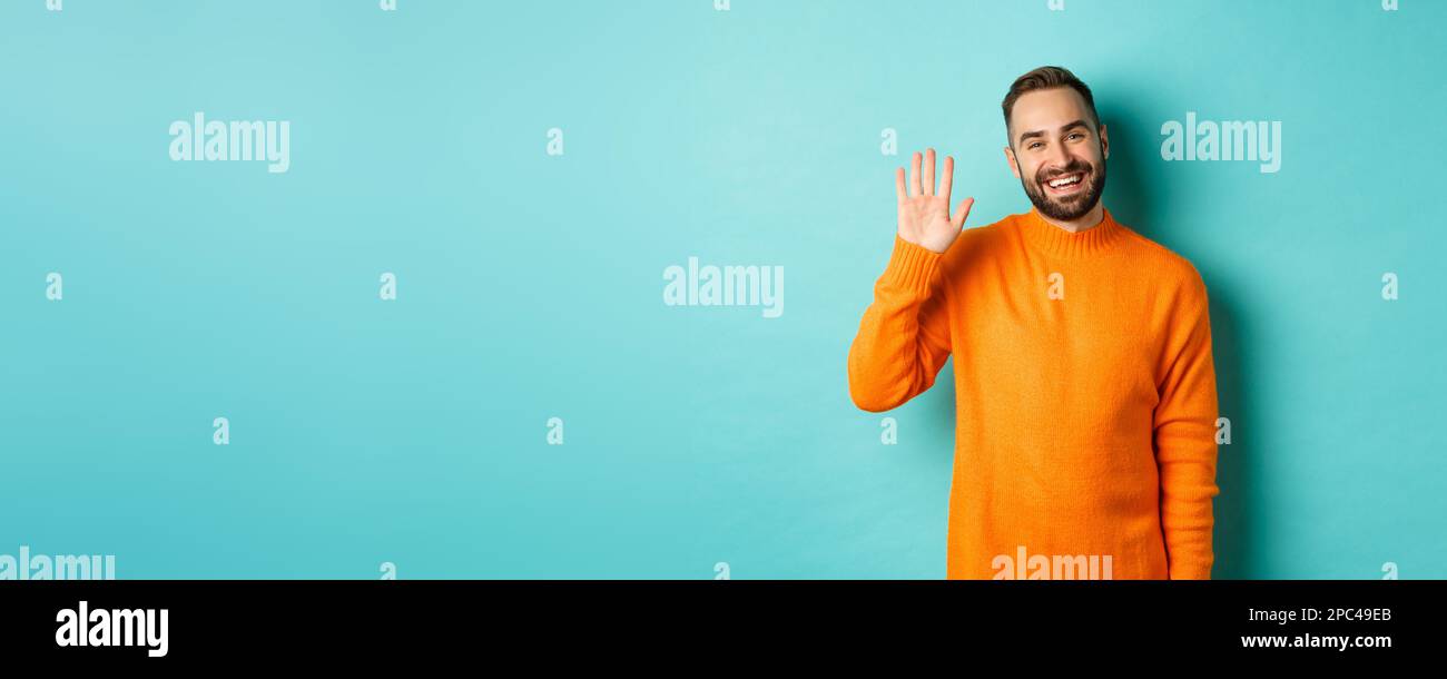 Photo of friendly young man saying hello, smiling and waiving hand ...
