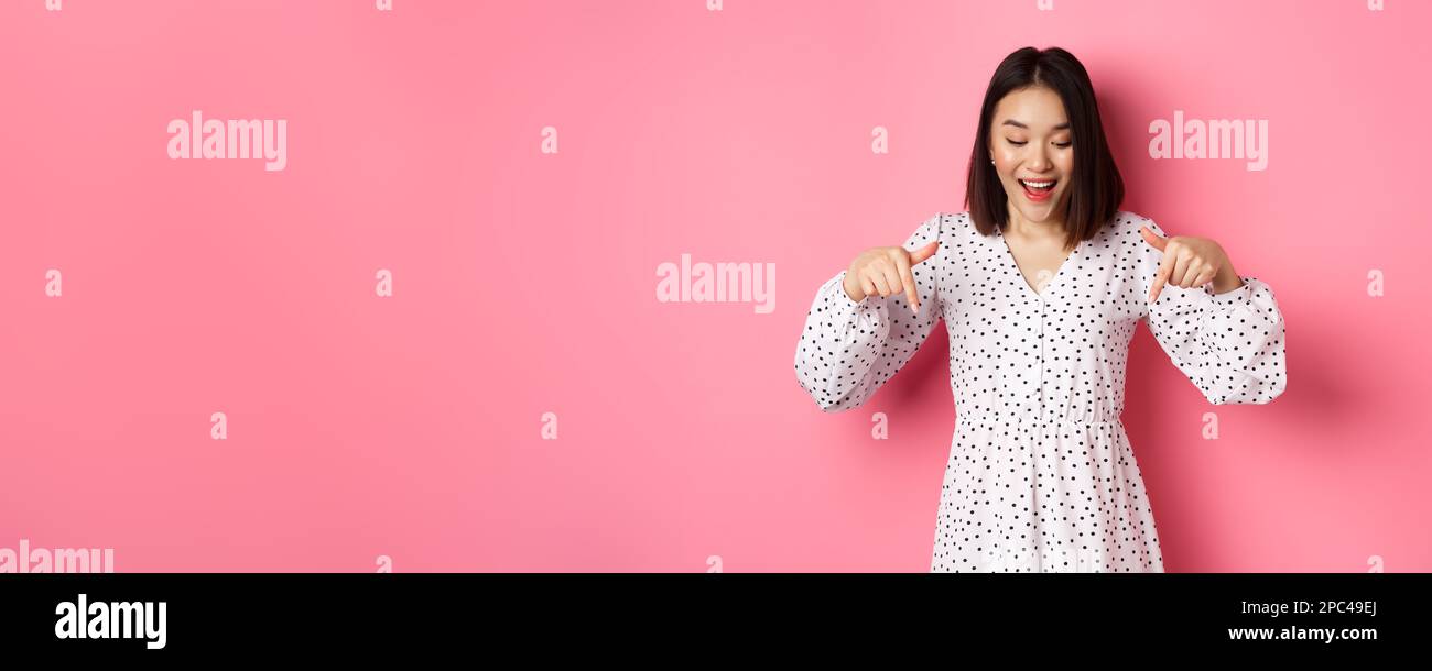 Romantic asian woman peeking down, pointing fingers at bottom, looking ...