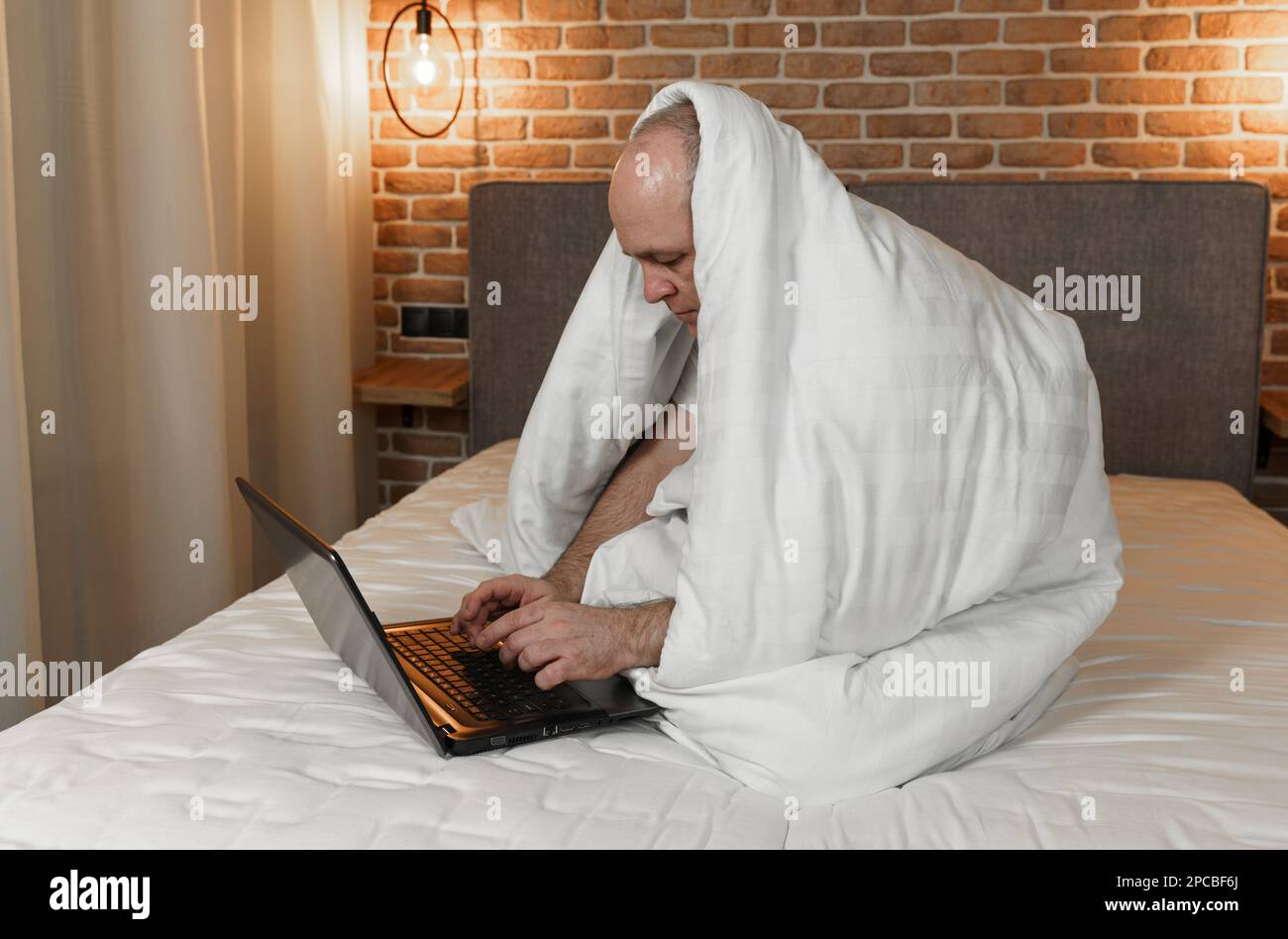 A man is sitting on a bed covered with a blanket, working at a computer ...