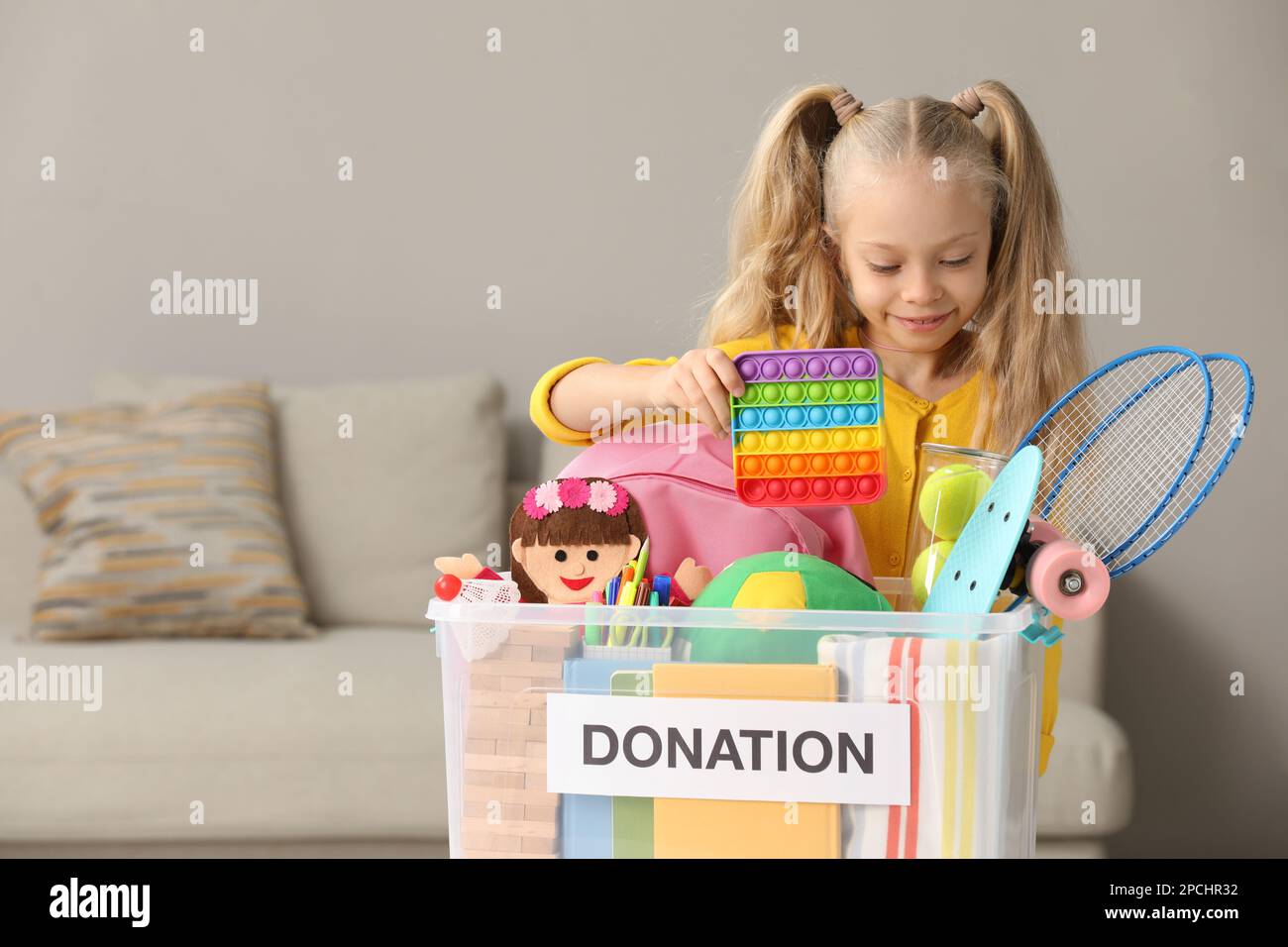 Cute little girl putting toy into donation box at home, space for text ...