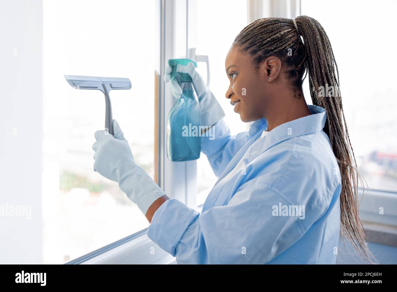 Cheerful young black woman housekeeper cleaning windows at apartment ...