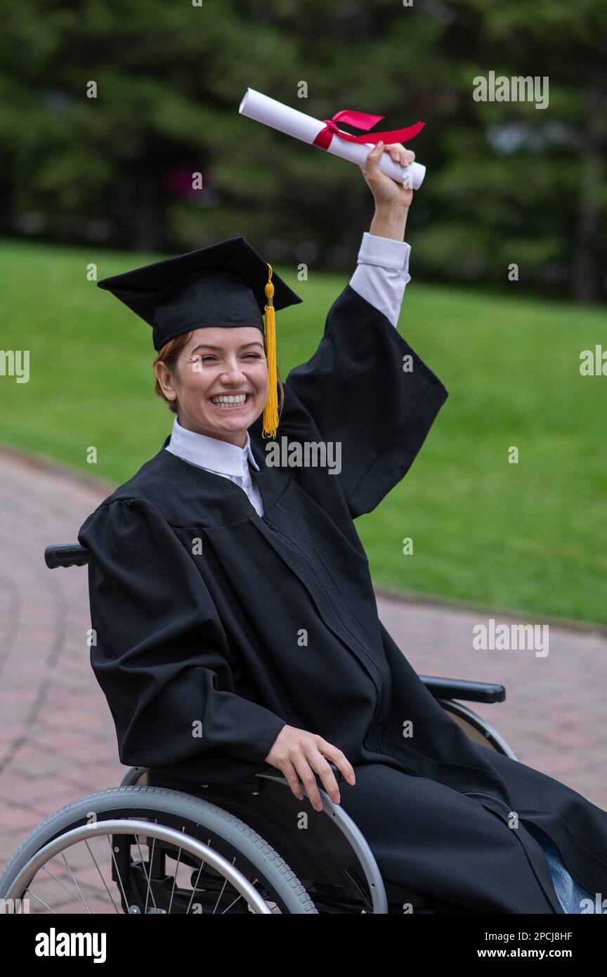 Caucasian woman in a wheelchair in a graduate costume rejoices at ...