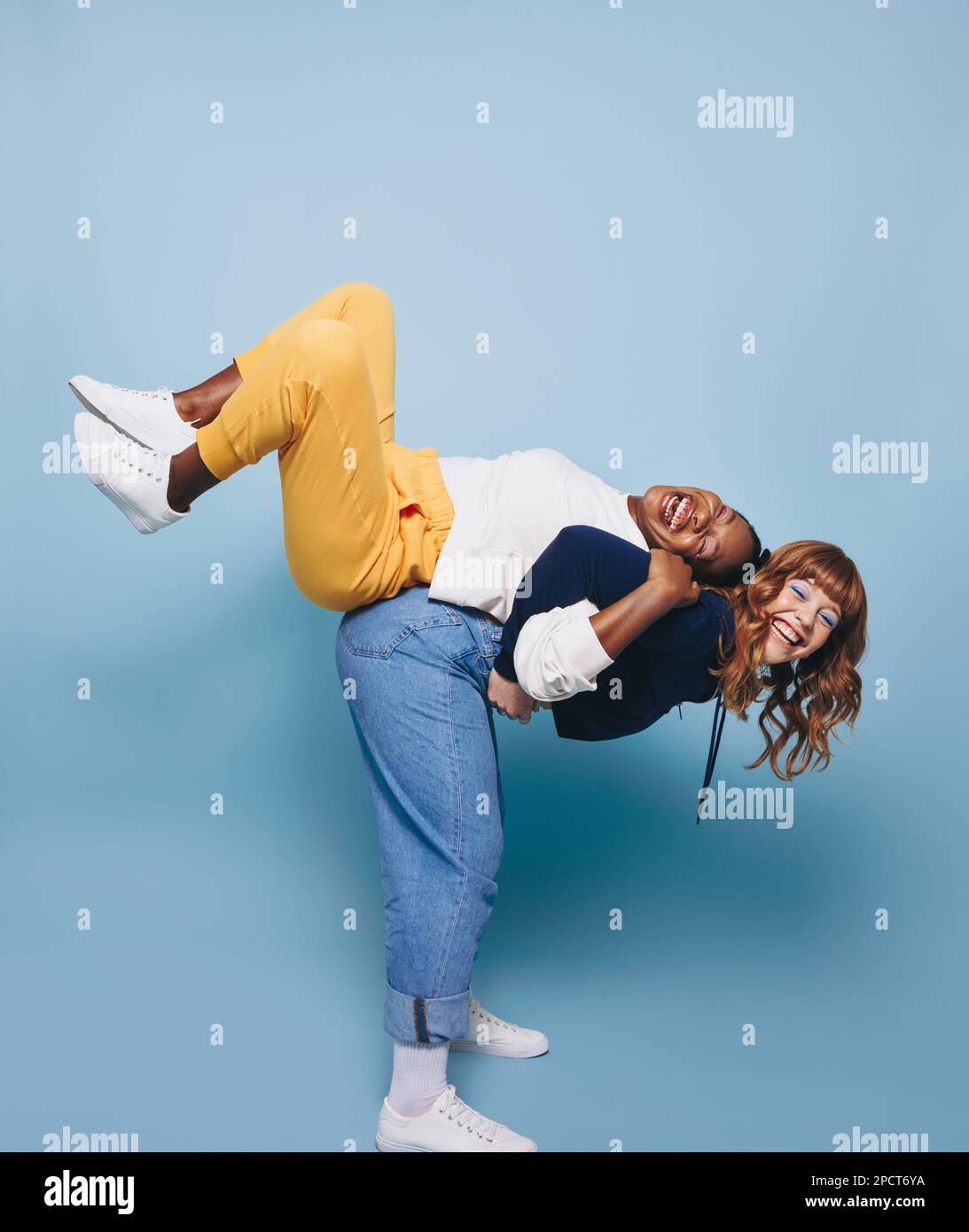 Playful young woman carrying her best friend on her back in a studio. Two vibrant young women ...