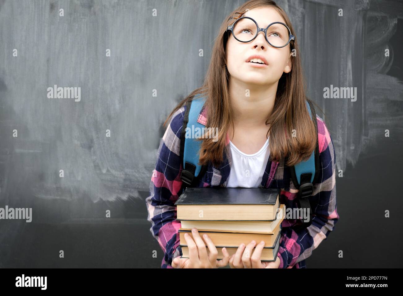 Teen girl little student holding pile of heavy books on chalkboard ...