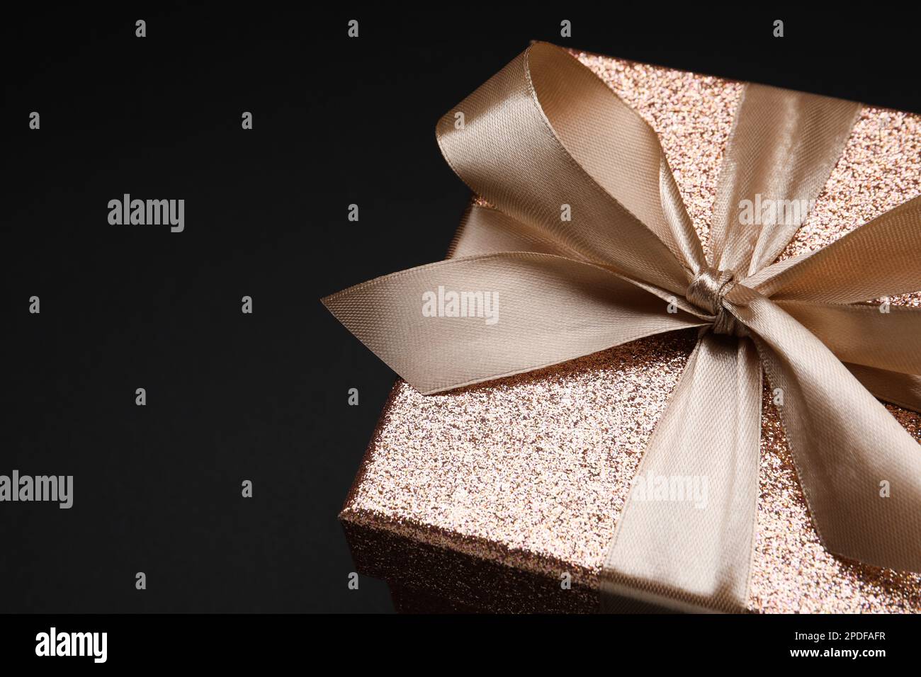 Shiny gift box with golden bow on black background, closeup. Space for ...