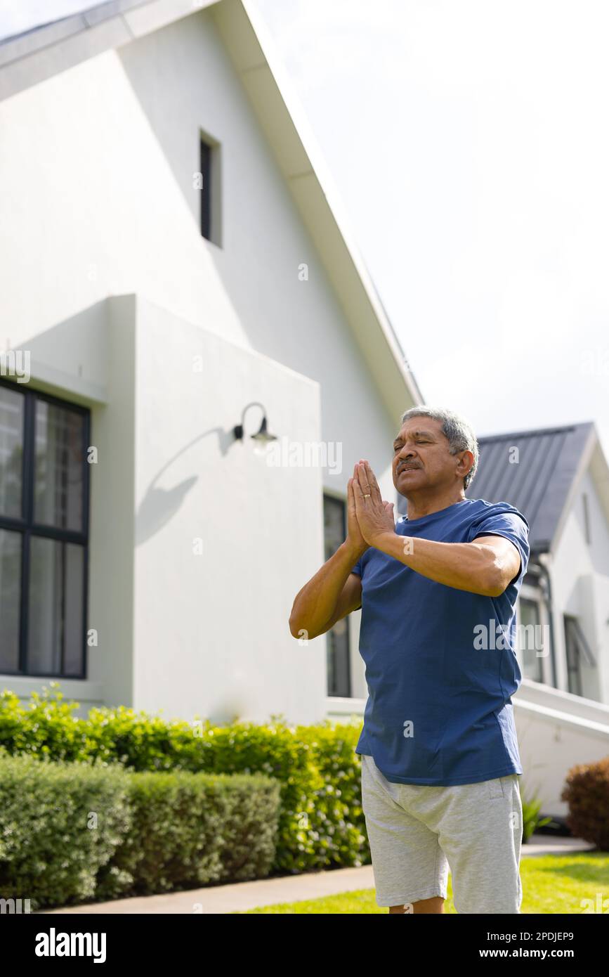 Low angle view of biracial senior man meditating in prayer pose while standing against house in ...