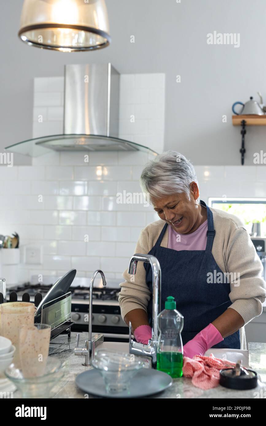 Smiling biracial senior woman wearing apron cleaning dishes in kitchen ...