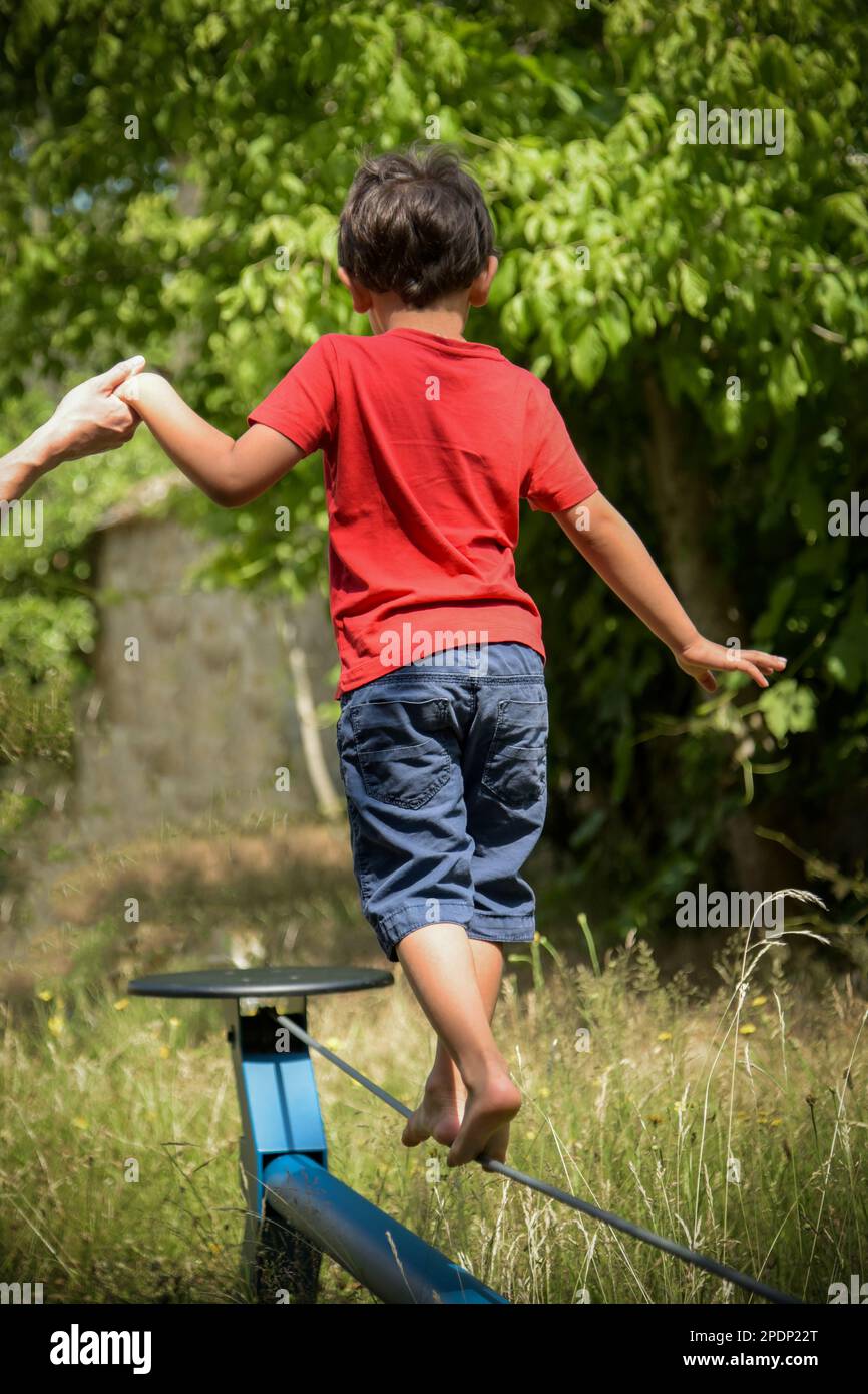 young boy walking on a wire with the help of a woman coach in a garden ...