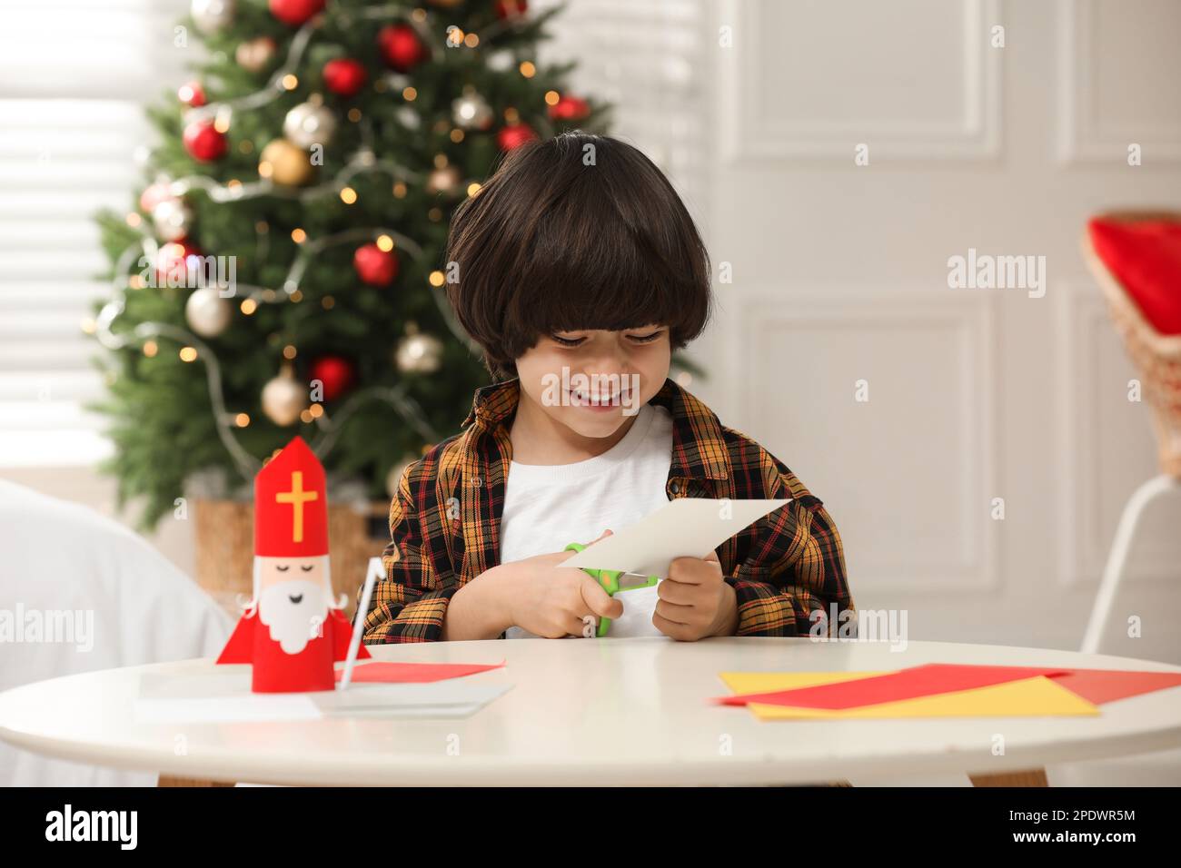 Cute little boy cutting paper at table with Saint Nicholas toy indoors ...