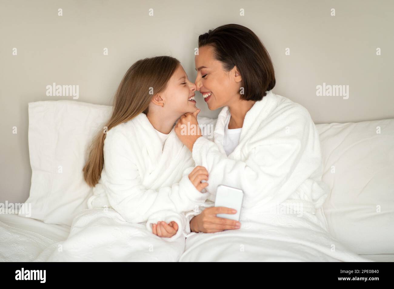Cheerful european adult woman and little girl in bathrobes rub noses ...