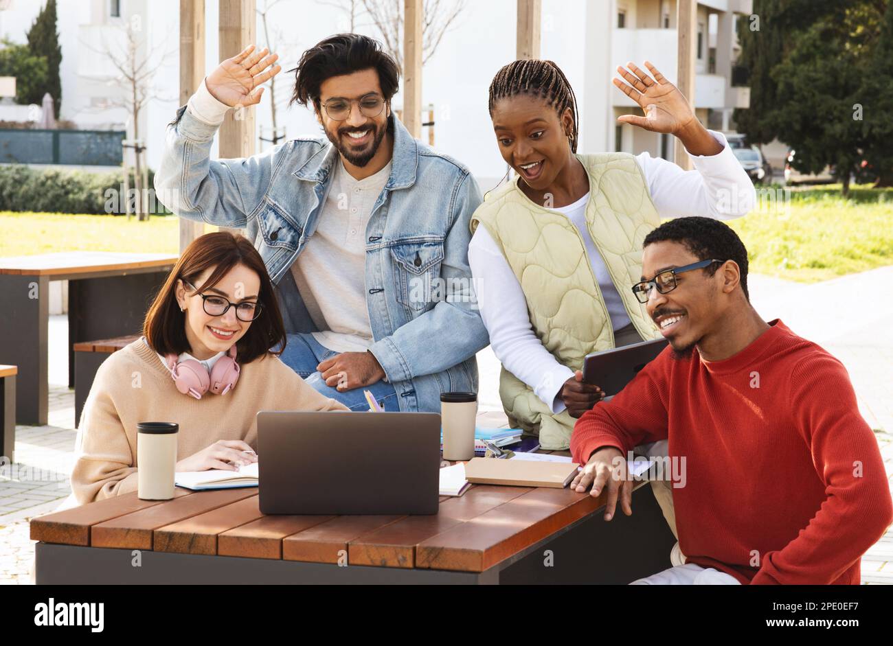 Smiling young multiethnic people students studying with laptop together, wave hand, say hi ...
