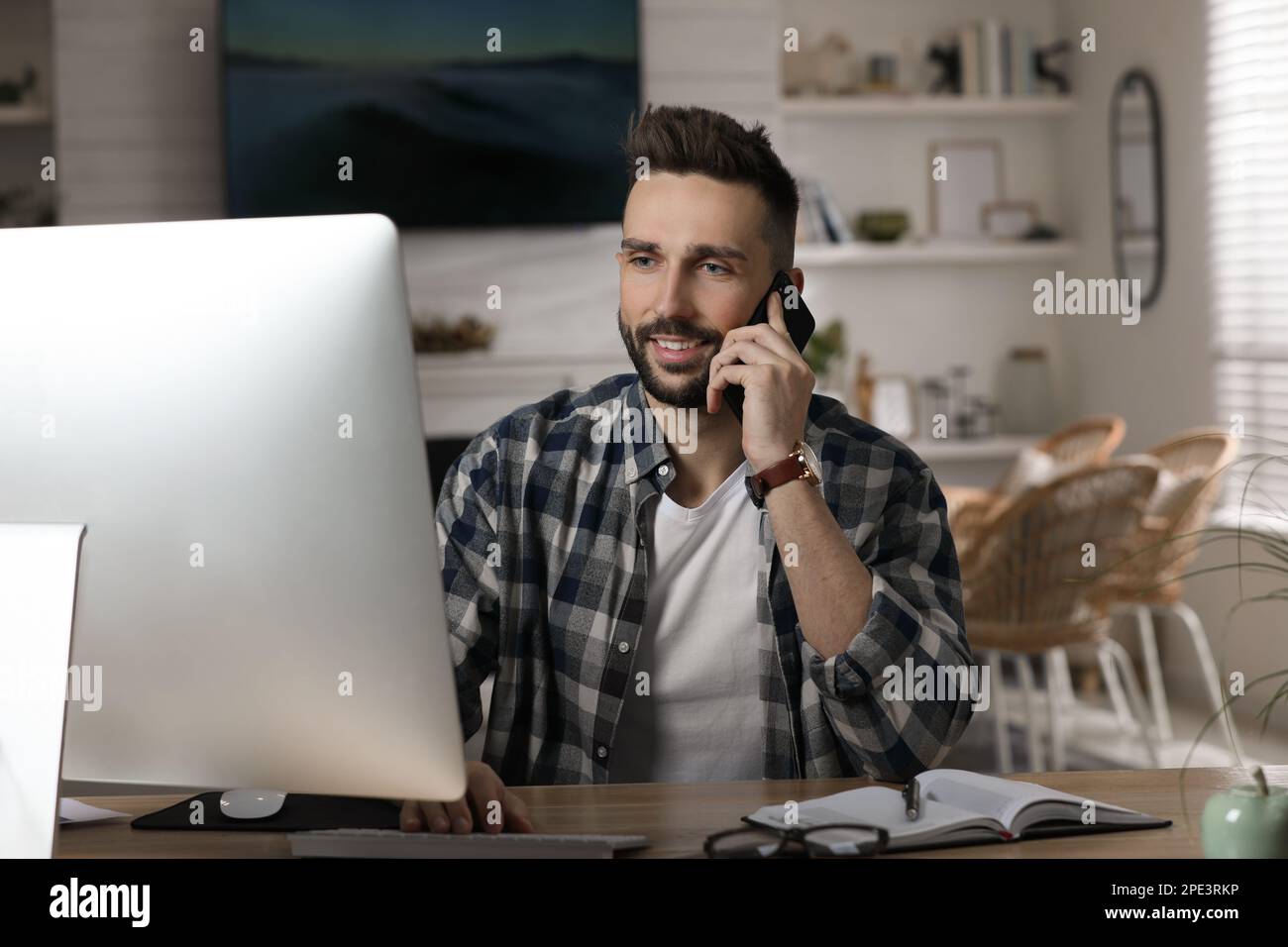 Man talking on phone while working with computer at table in home ...