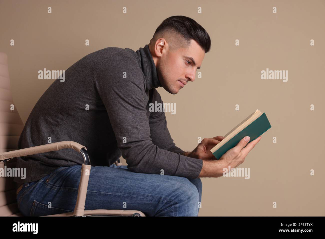 Man with poor posture reading book while sitting on chair against beige ...