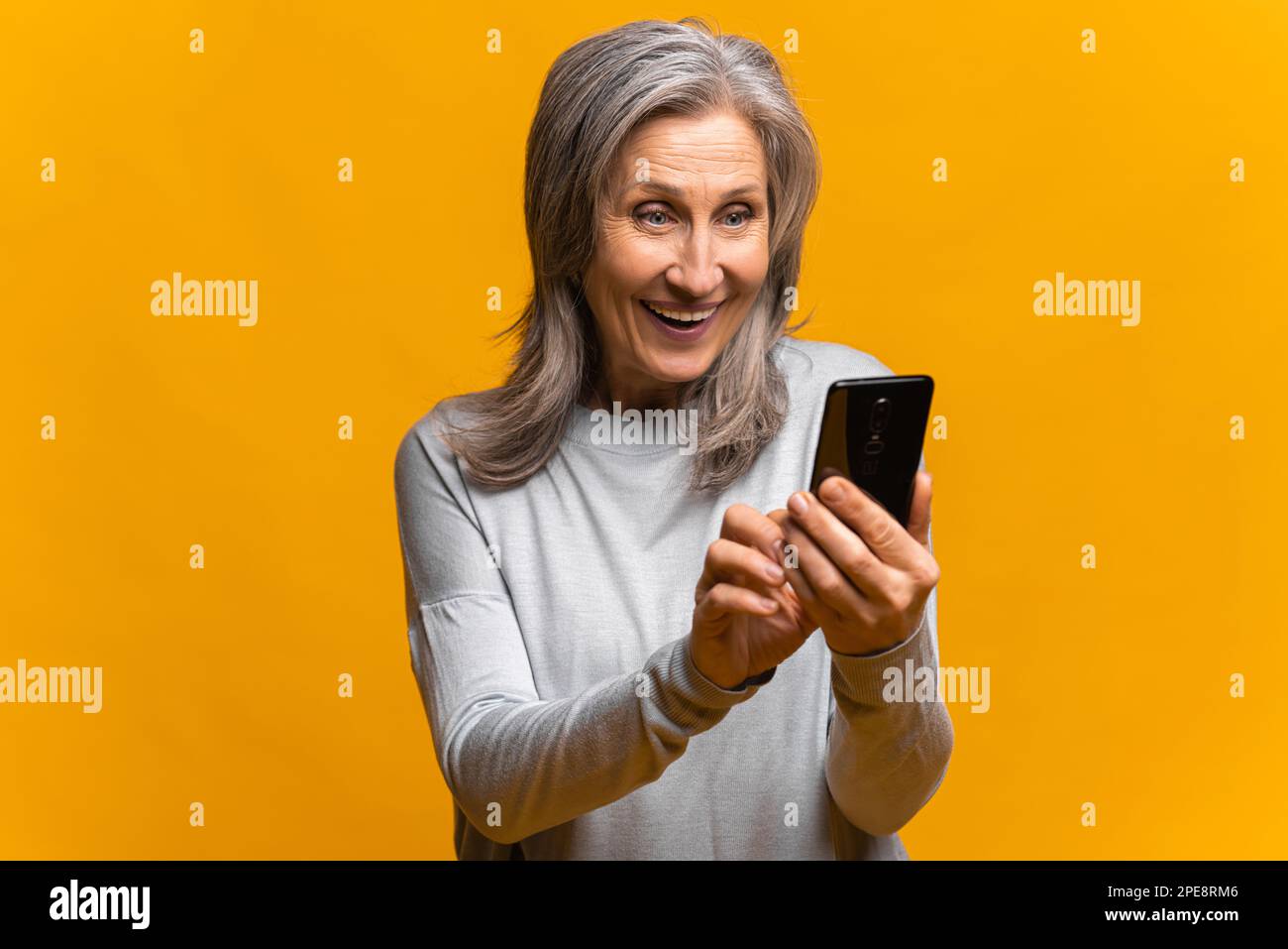 Shocked senior woman standing, holding and pointing at mobile display ...