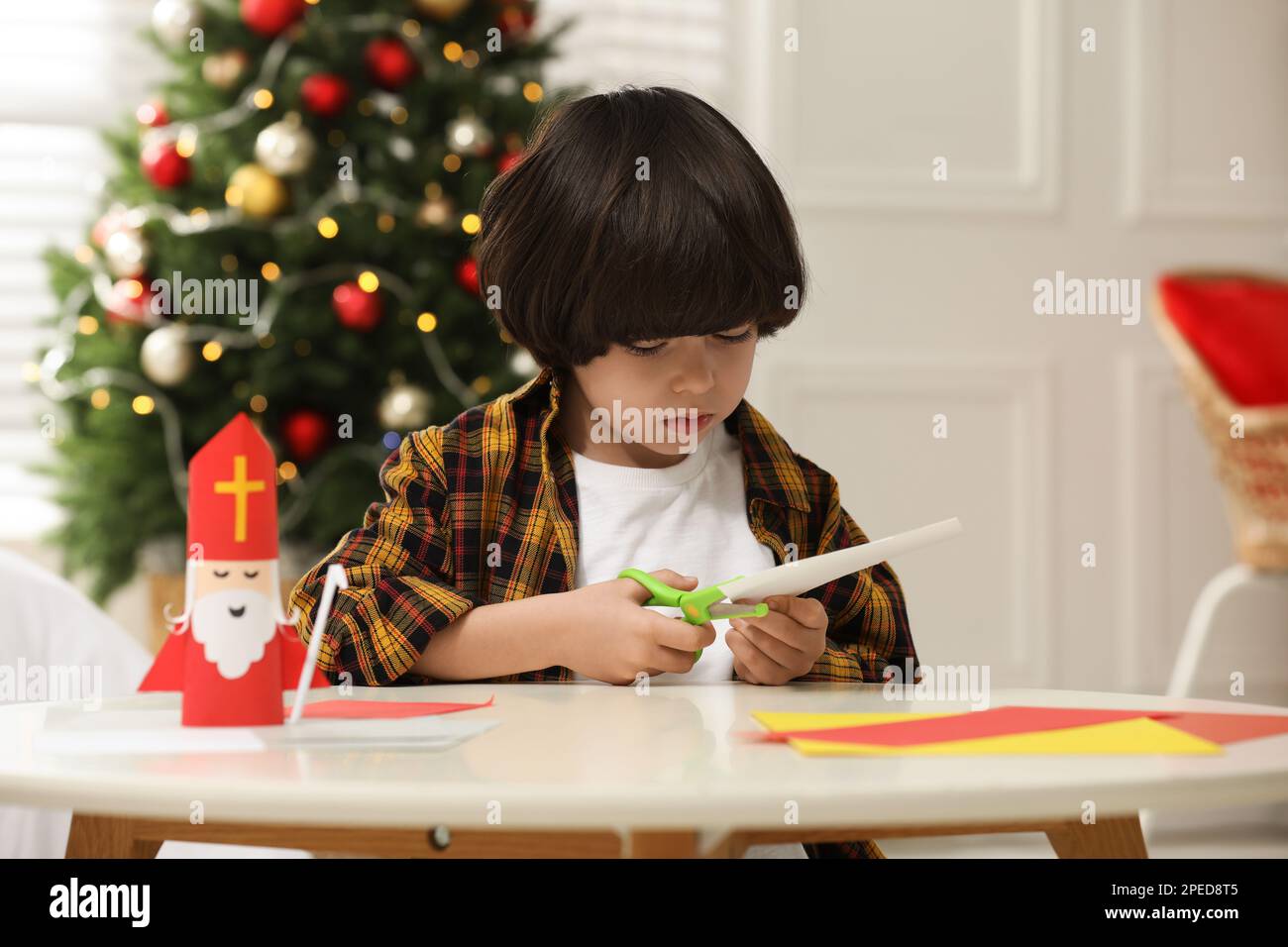 Cute little boy cutting paper at table with Saint Nicholas toy indoors ...