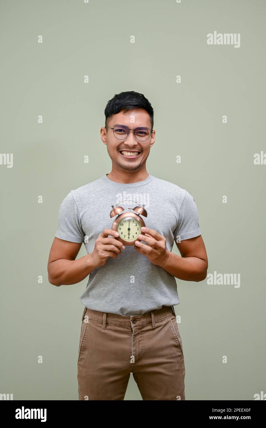 Smiling millennial Asian man in casual clothes holding a vintage alarm ...