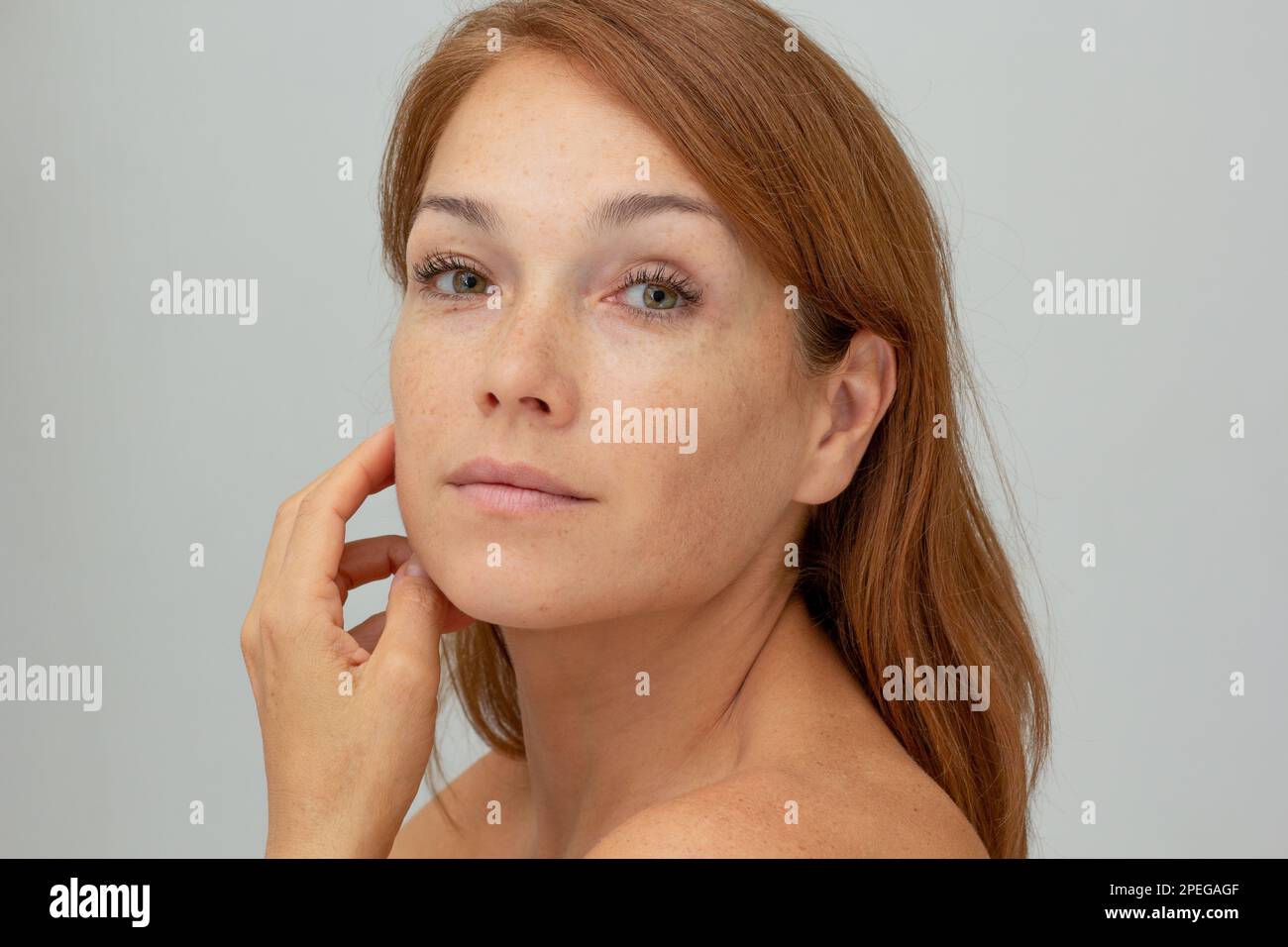 Portrait of middle aged woman with reddish hair holding hand on cheek looking at camera on grey ...