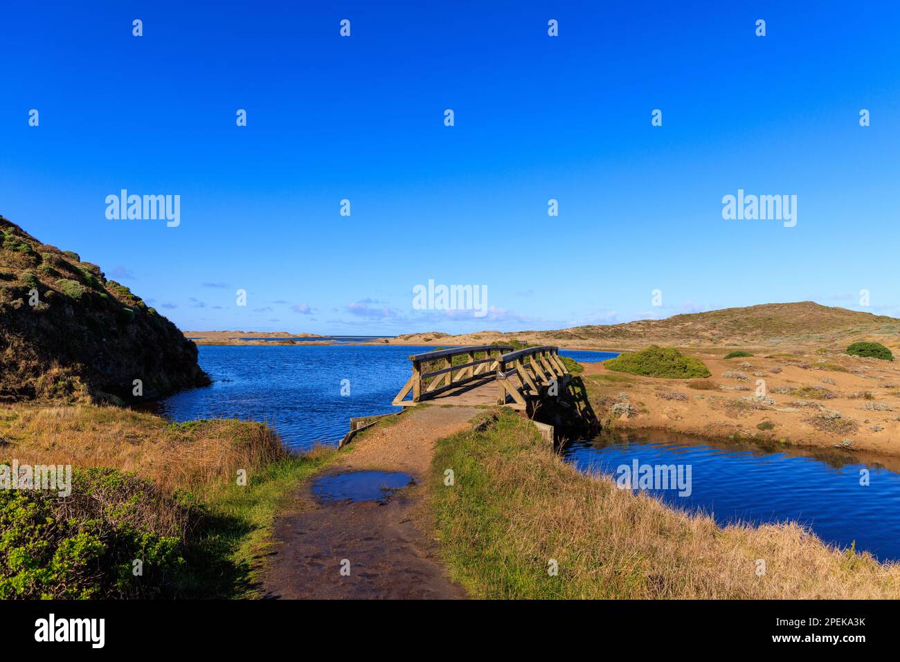 Wooden footbridge over blue water along dirt trail in dry coastal ...