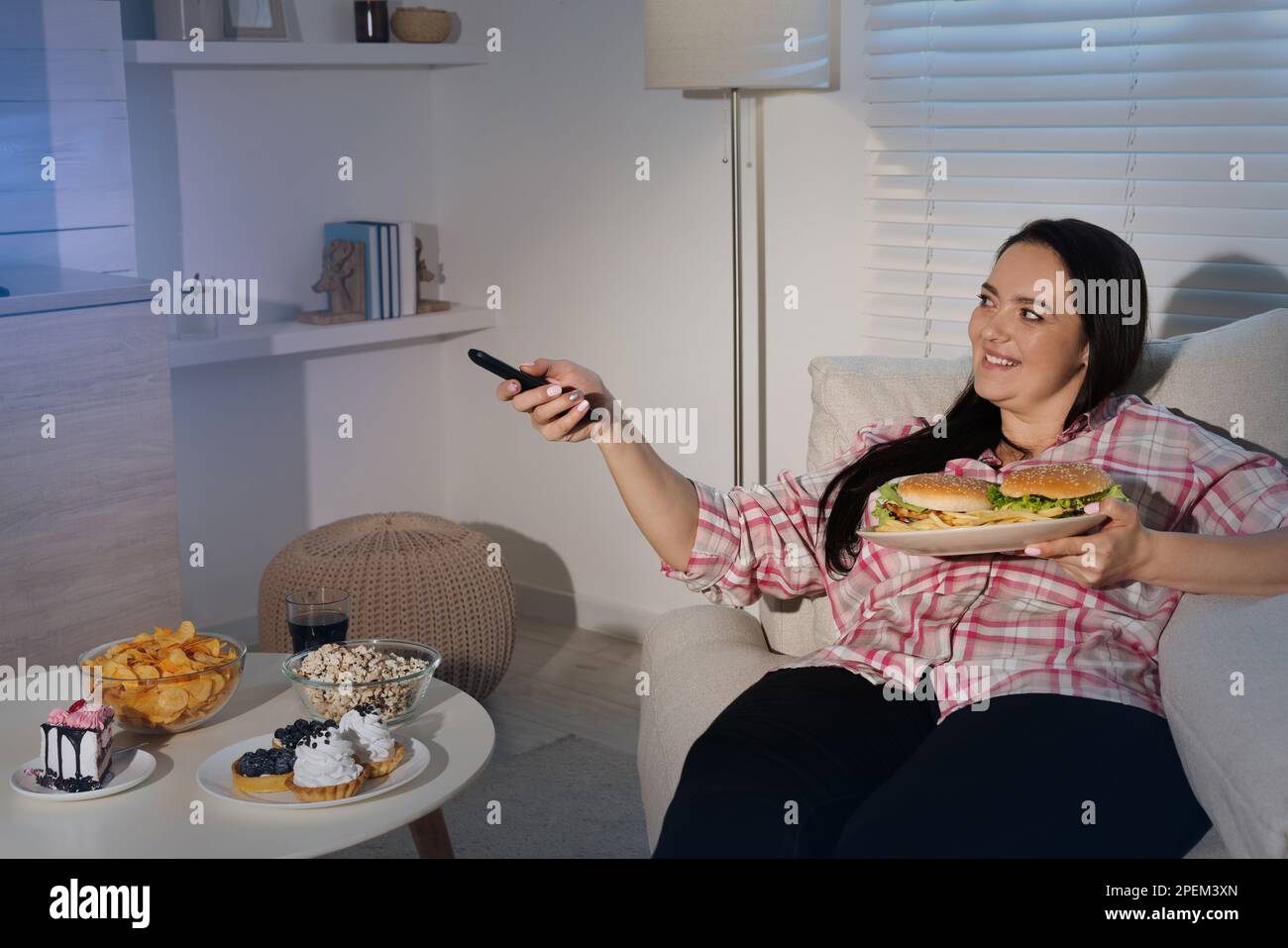 Happy overweight woman with unhealthy food watching TV on sofa at home ...