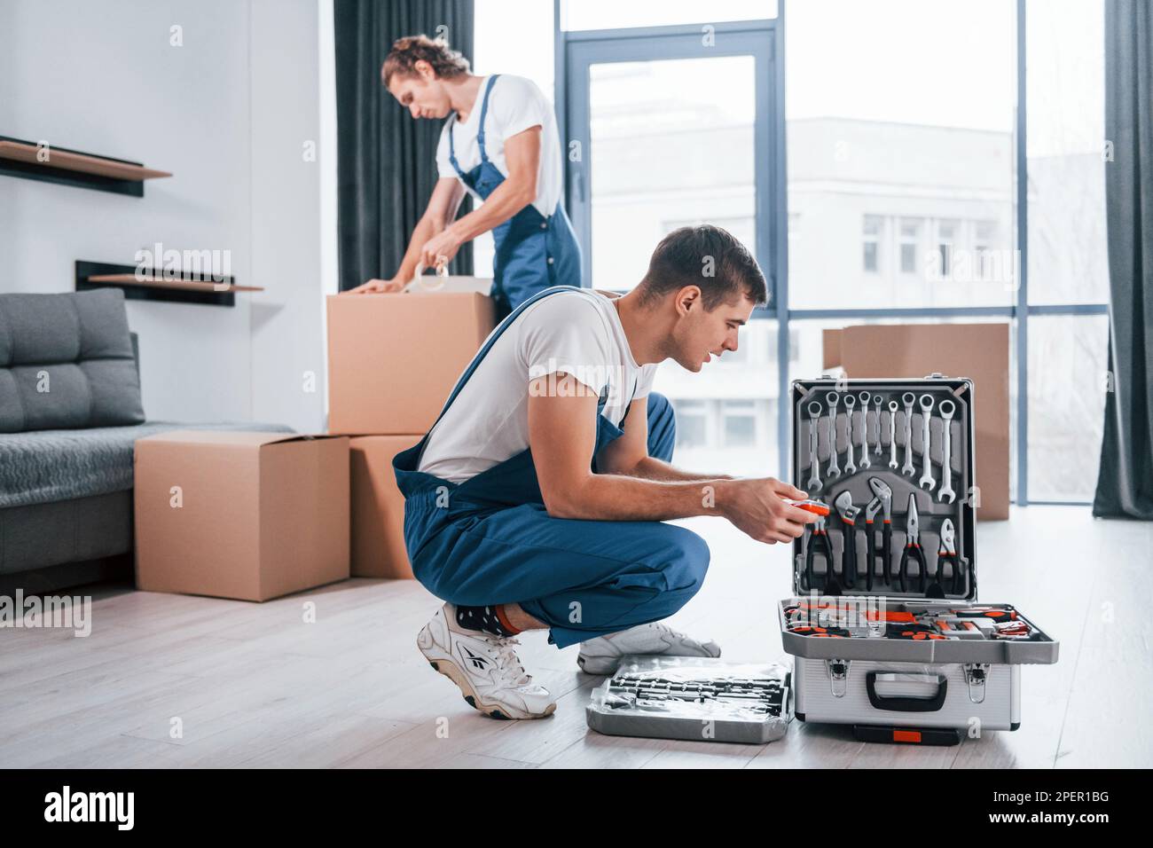 Uses tools from case. Two young movers in blue uniform working indoors ...