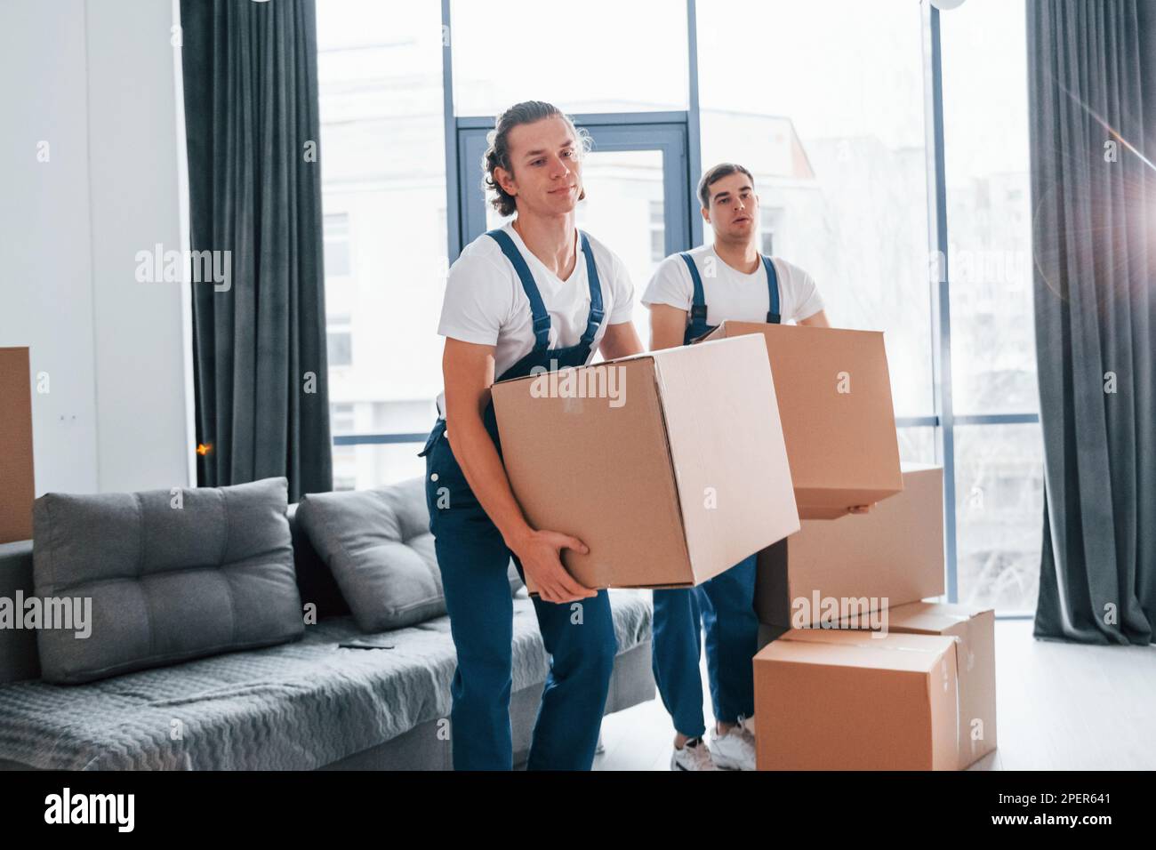 Busy day. Two young movers in blue uniform working indoors in the room ...