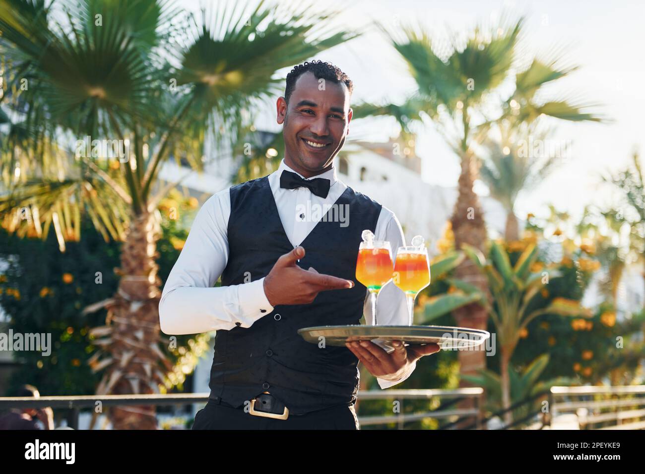 Holds cocktails. Black waiter in formal clothes is at his work outdoors ...