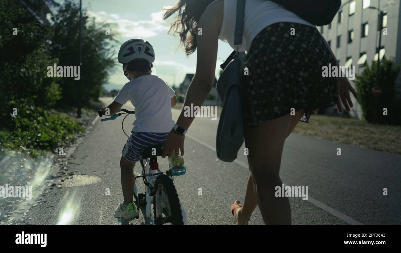 Back of child learning to ride bicycle outside with mother help and ...