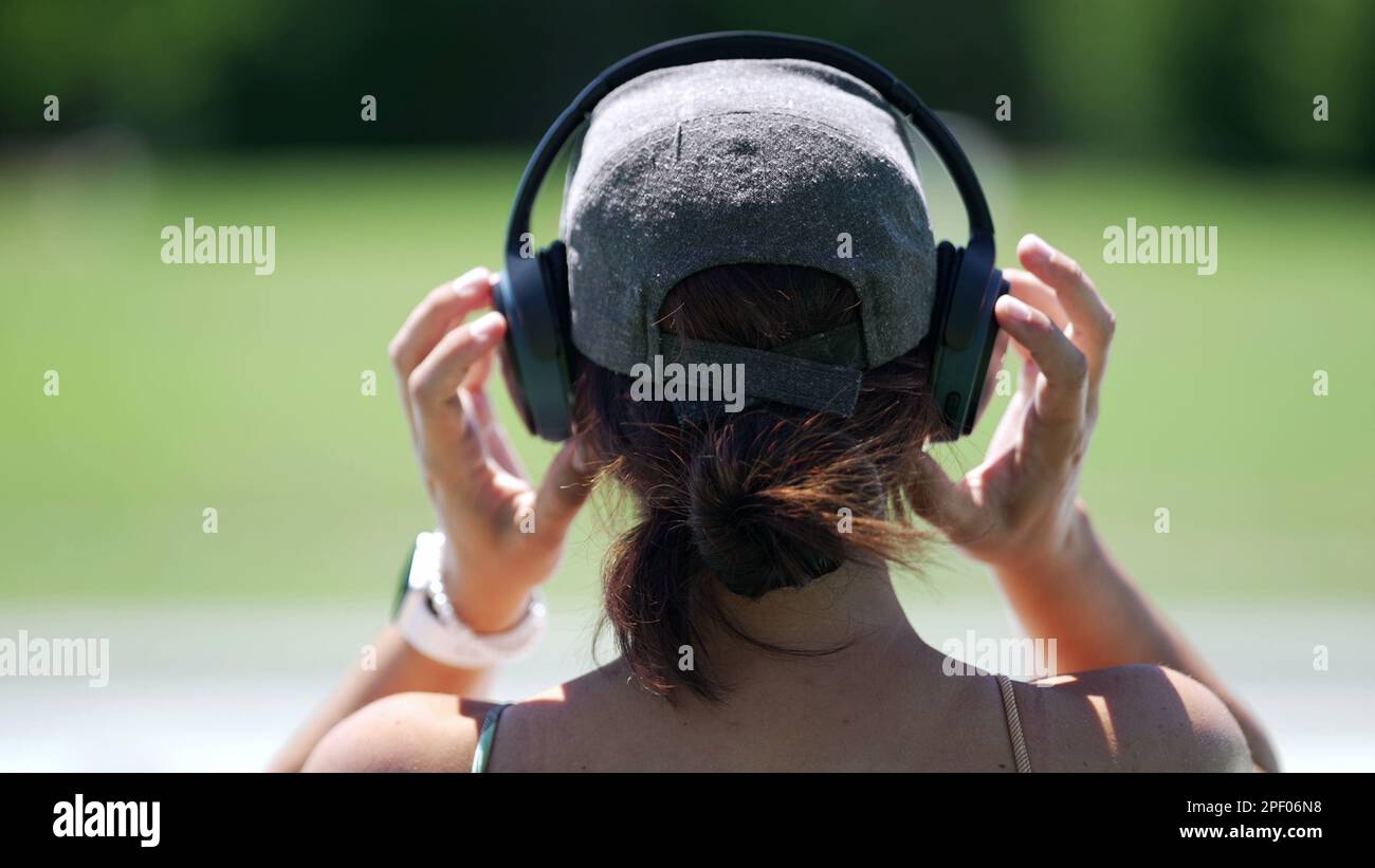 Back of person removing headphones sitting outside at park bench. Woman ...