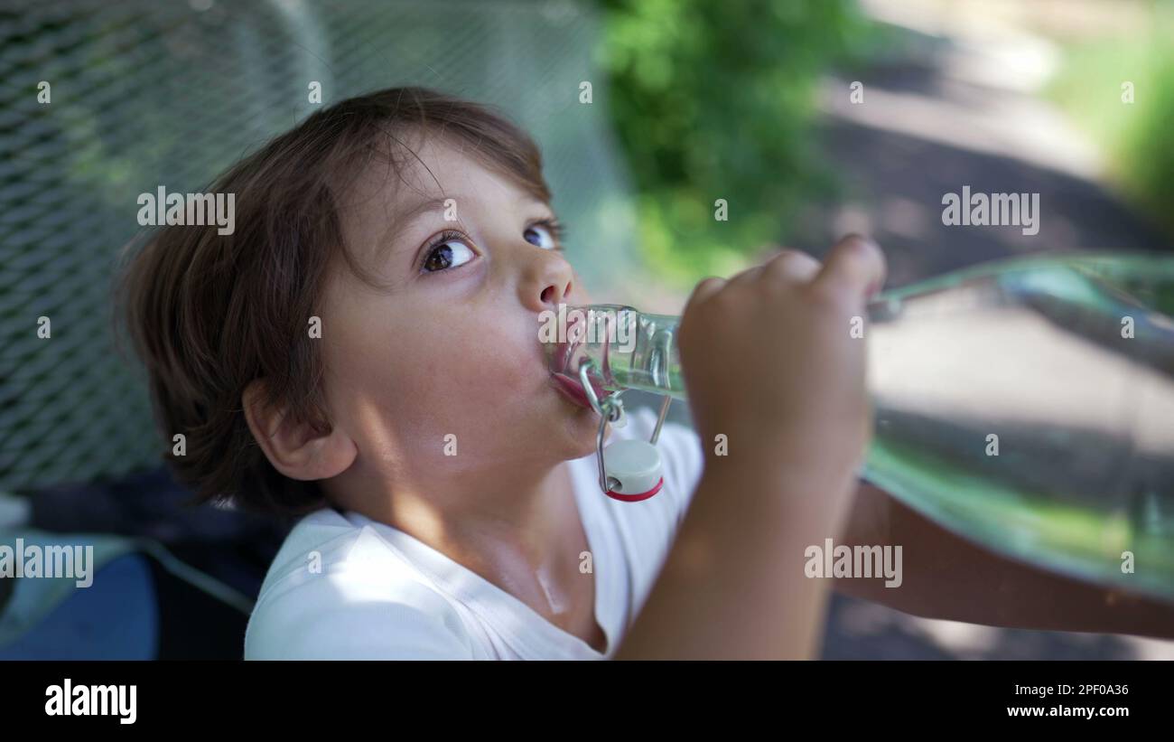 Child drinks water during hot summer day. Little boy gulping from glass bottle hydrating liquid ...