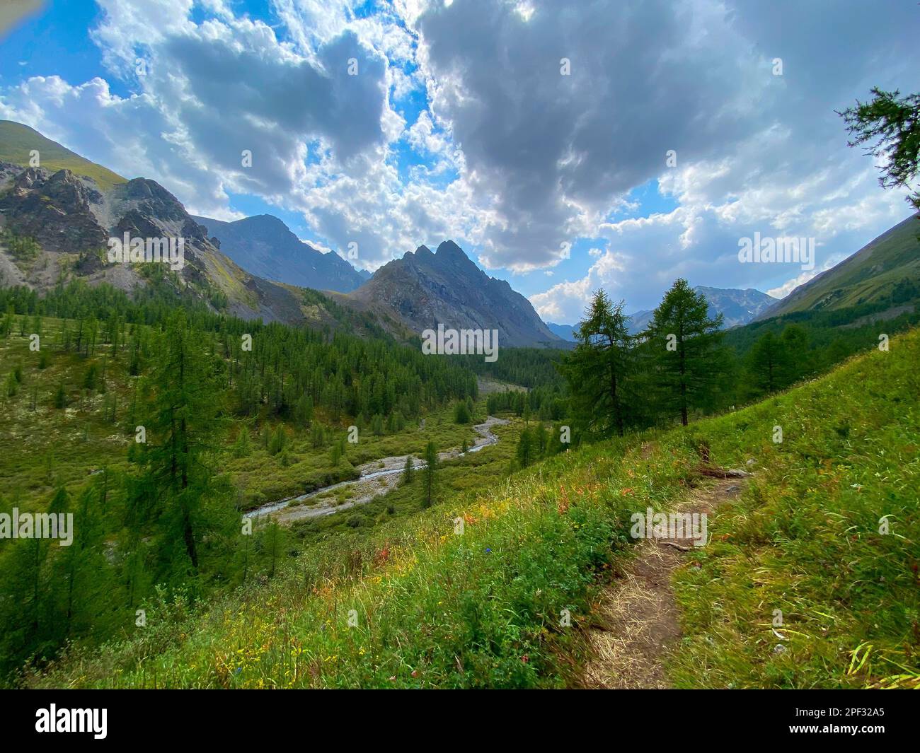 Panorama trail in grass field between mountains and rocks under clouds ...