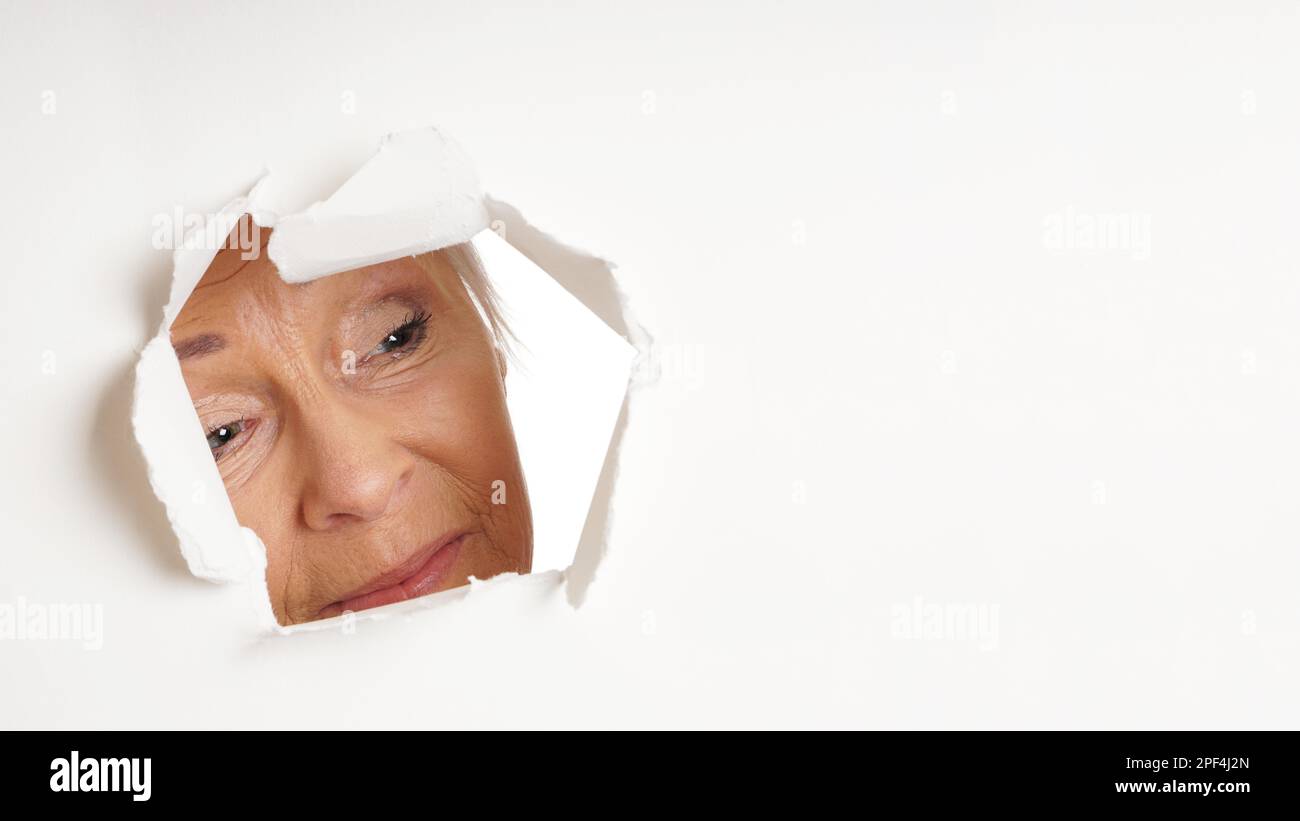 curious older woman looking through hole torn in white paper background ...