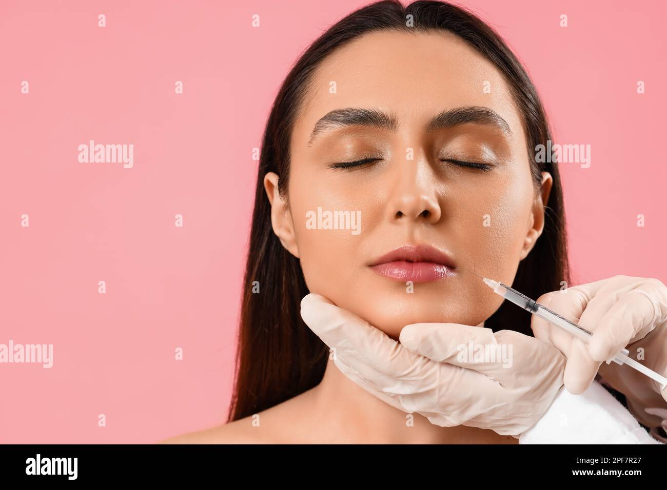 Young woman receiving filler injection in face against pink background ...