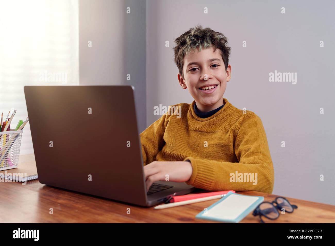 A smiling 10-year-old child is sitting at a desk in front of a laptop ...
