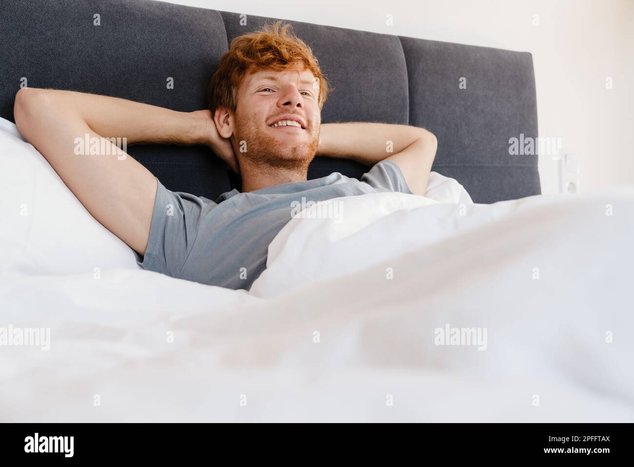 Young handsome smiling redhead man in gray t-shirt with hands behind ...
