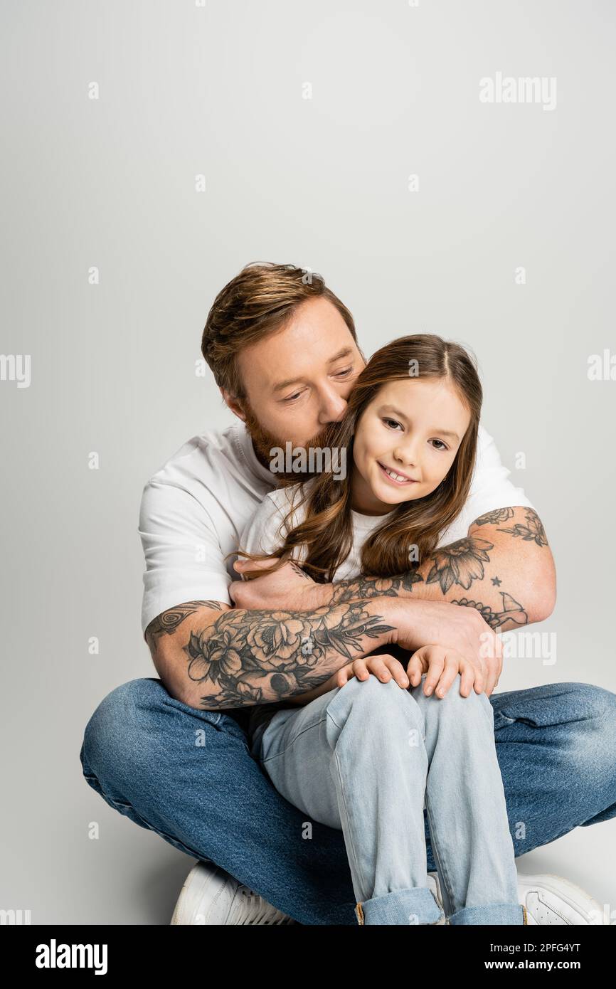 Tattooed man hugging and kissing head of happy daughter on grey ...