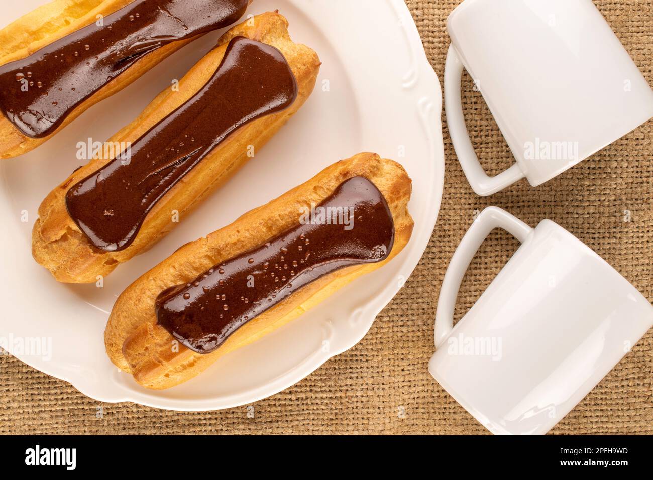 Three chocolate eclairs with a white ceramic plate and two cups on a ...