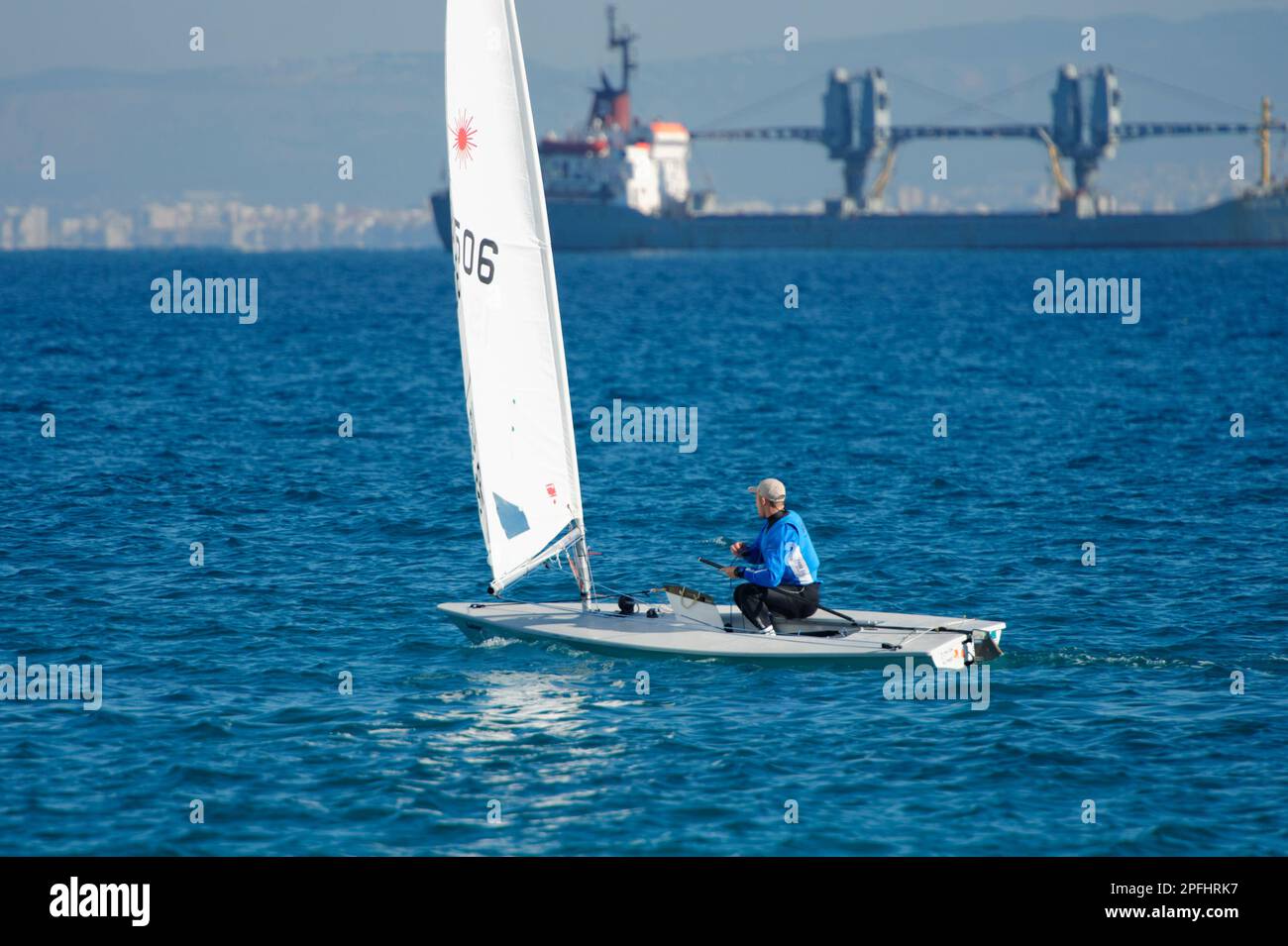 Photo of a teenager sailing a laser class sailboat with a huge ship on ...