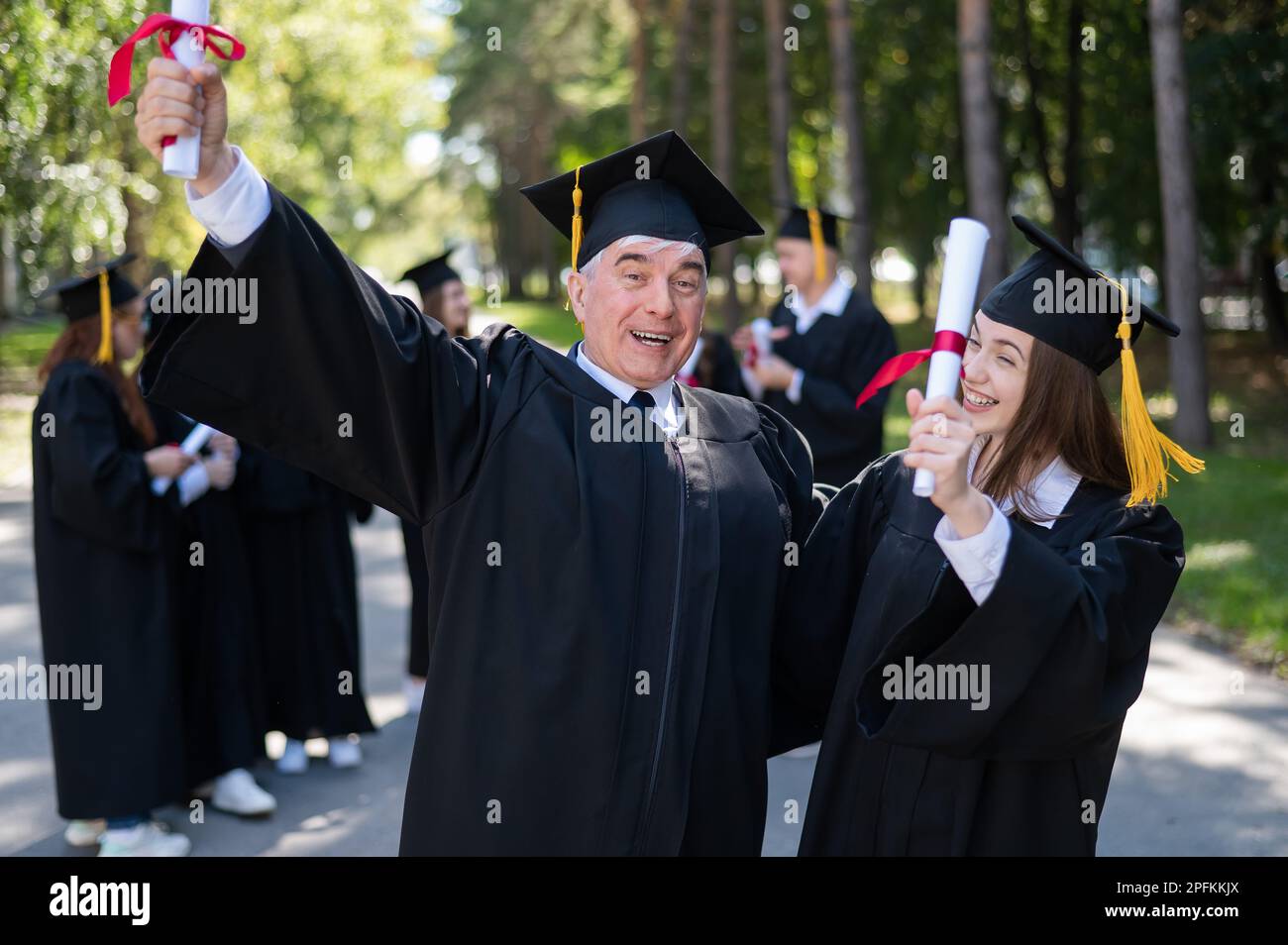 A group of graduates in robes outdoors. An elderly man and a young ...