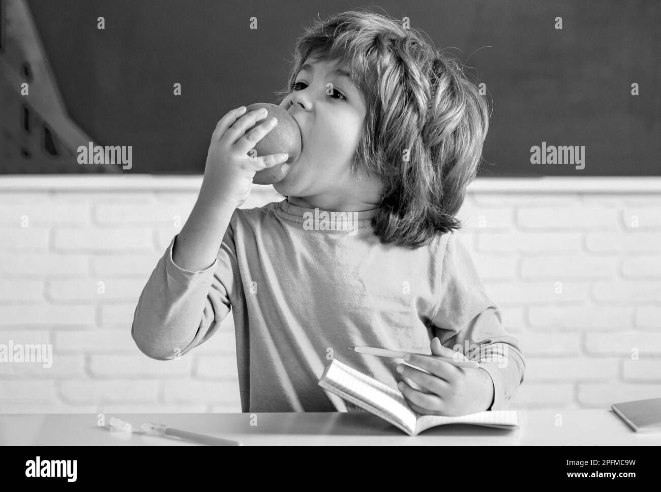 Friendly child eat apple in classroom near blackboard desk. Elementary ...