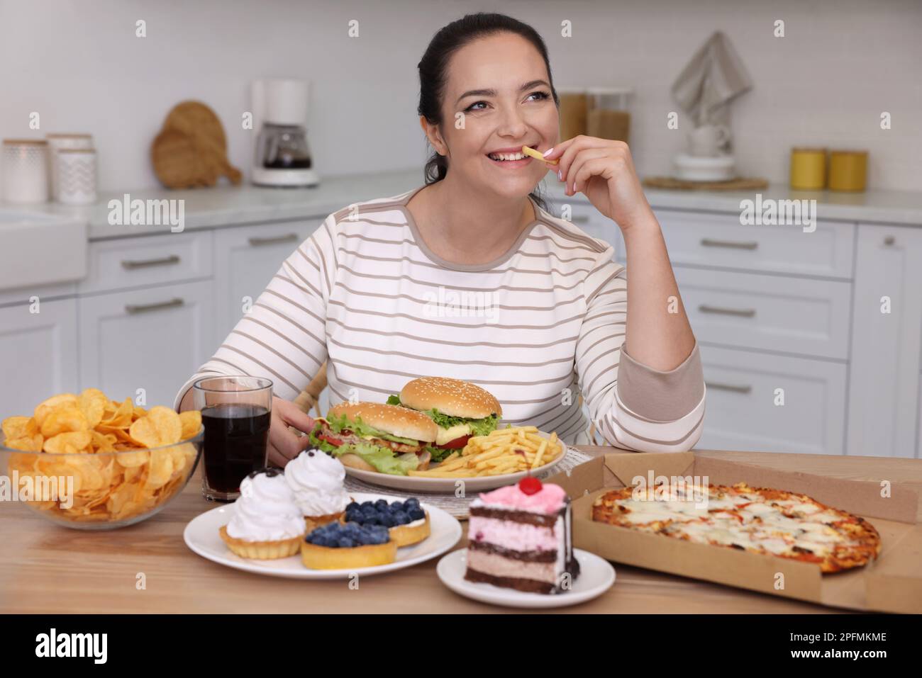 Happy overweight woman eating French fries at table ladened with other ...