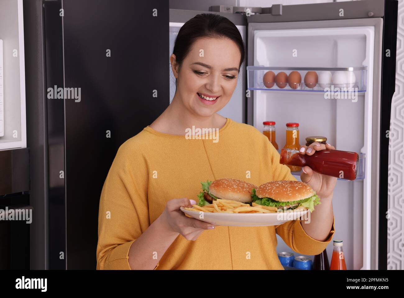 Happy Overweight Woman With Ketchup And Burgers Near Fridge In Kitchen