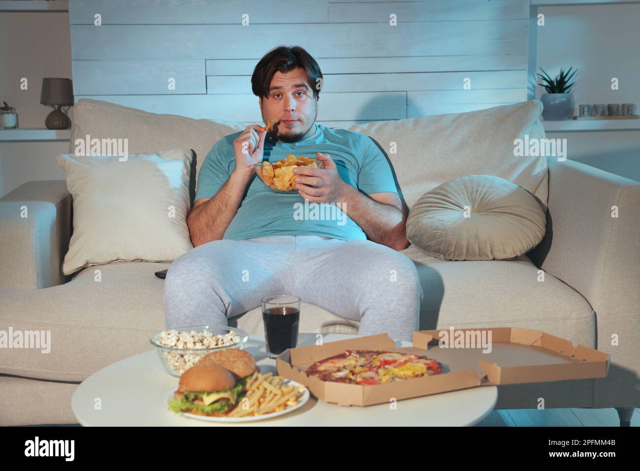 Overweight man with chips watching TV on sofa at home Stock Photo - Alamy