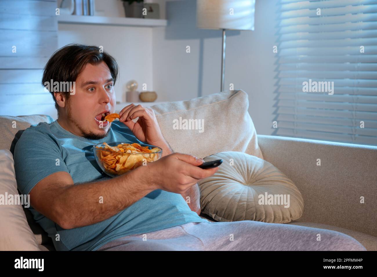 Overweight man with chips watching TV on sofa at home Stock Photo - Alamy