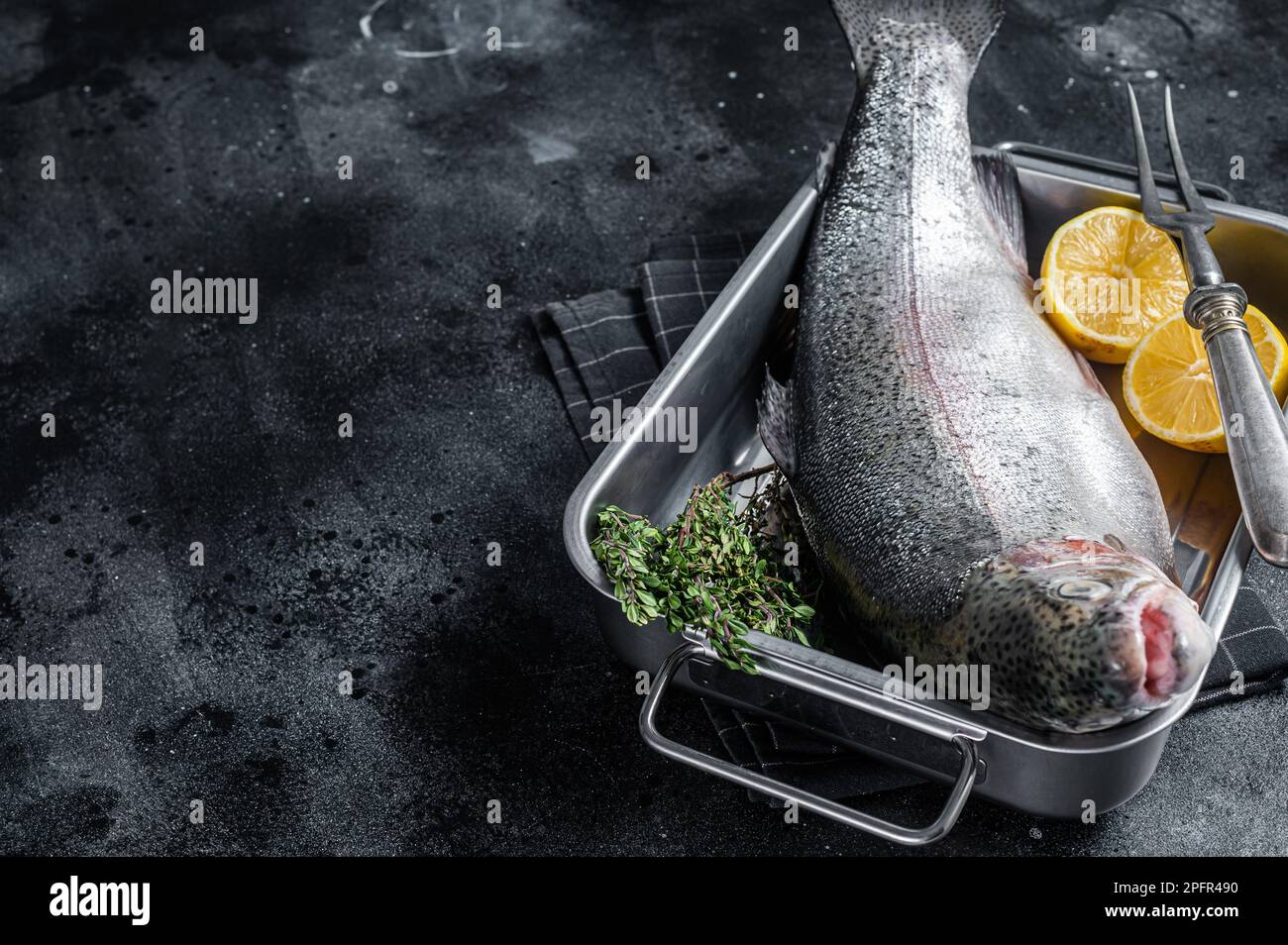 Raw whole trout fish in a kitchen tray with herbs. Black background ...